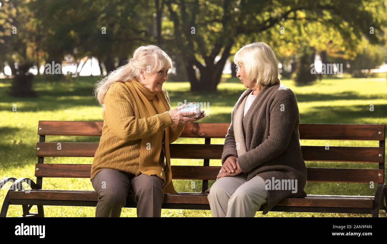 Old woman giving birthday present to friend on bench in park, golden ...