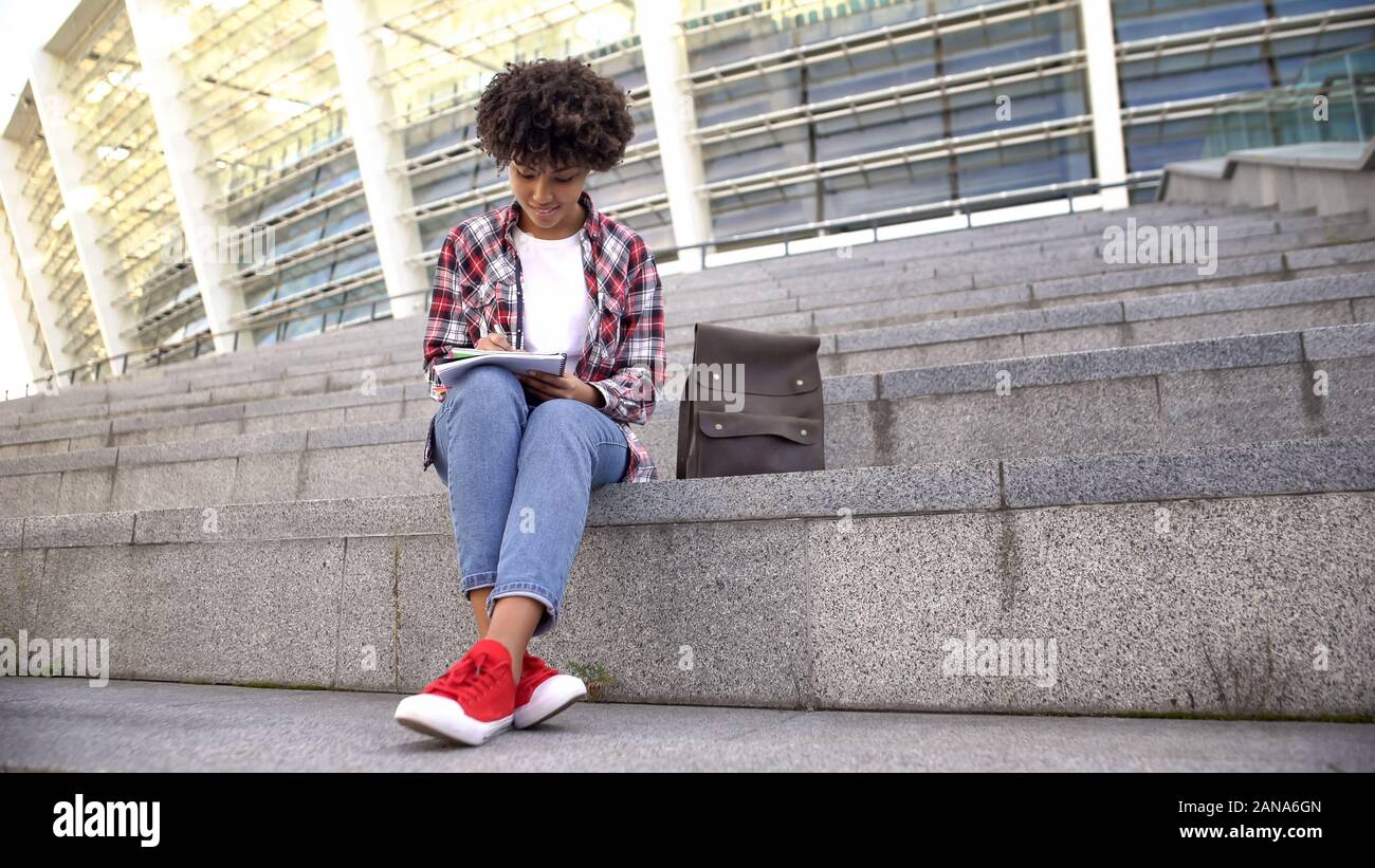 Smart afro-american student writing in notebook, doing homework near ...