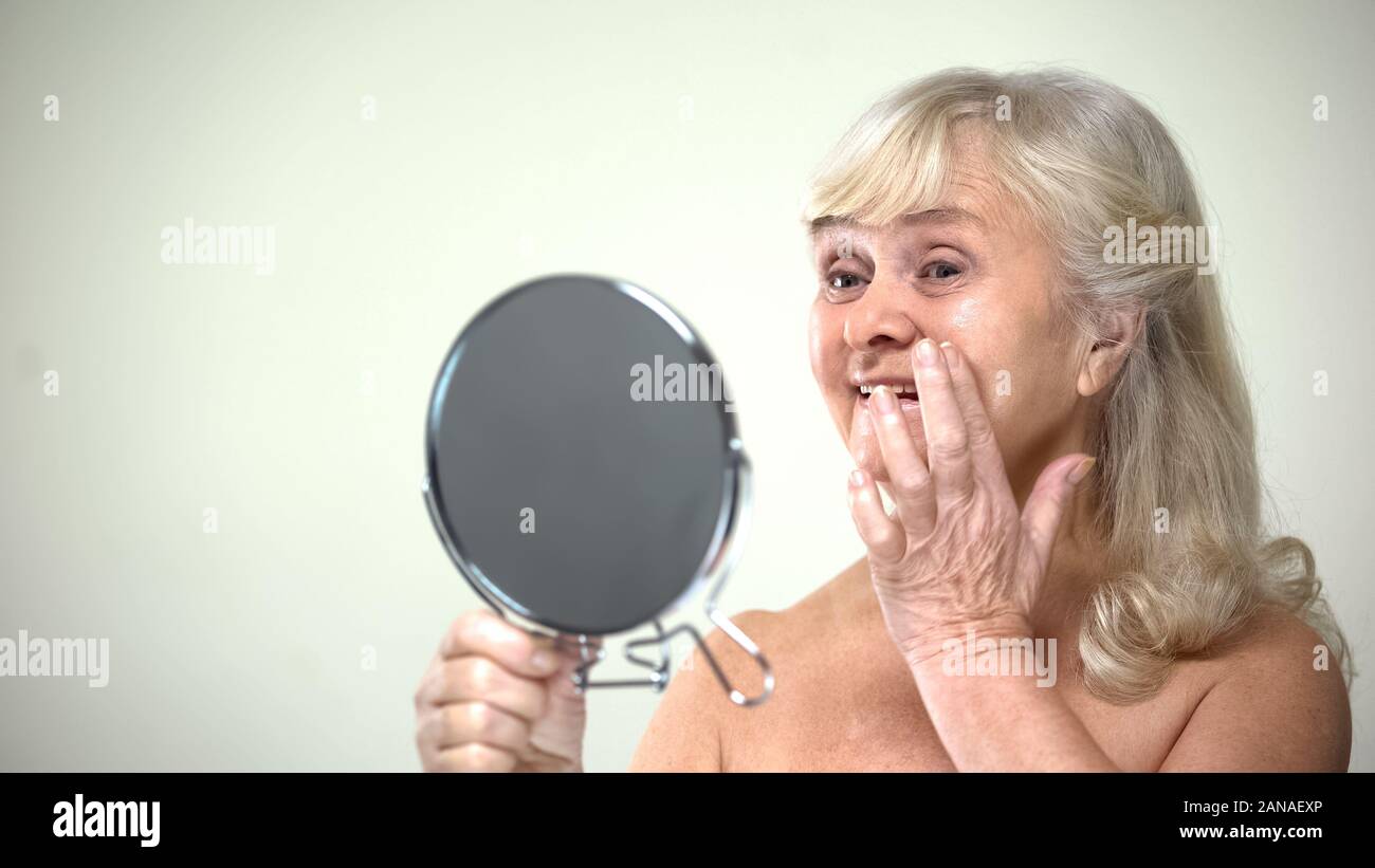 Optimistic elderly lady applying anti-aging cream looking in mirror ...