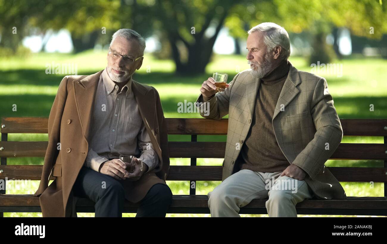 Serious gentlemen drinking whiskey, sitting on bench in park ...