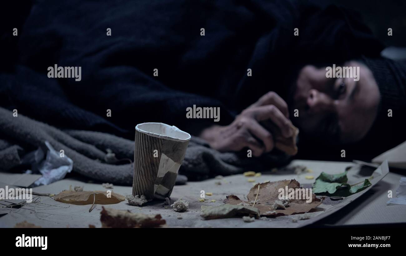 Homeless person lying on street eating last piece of bread, paper cup ...