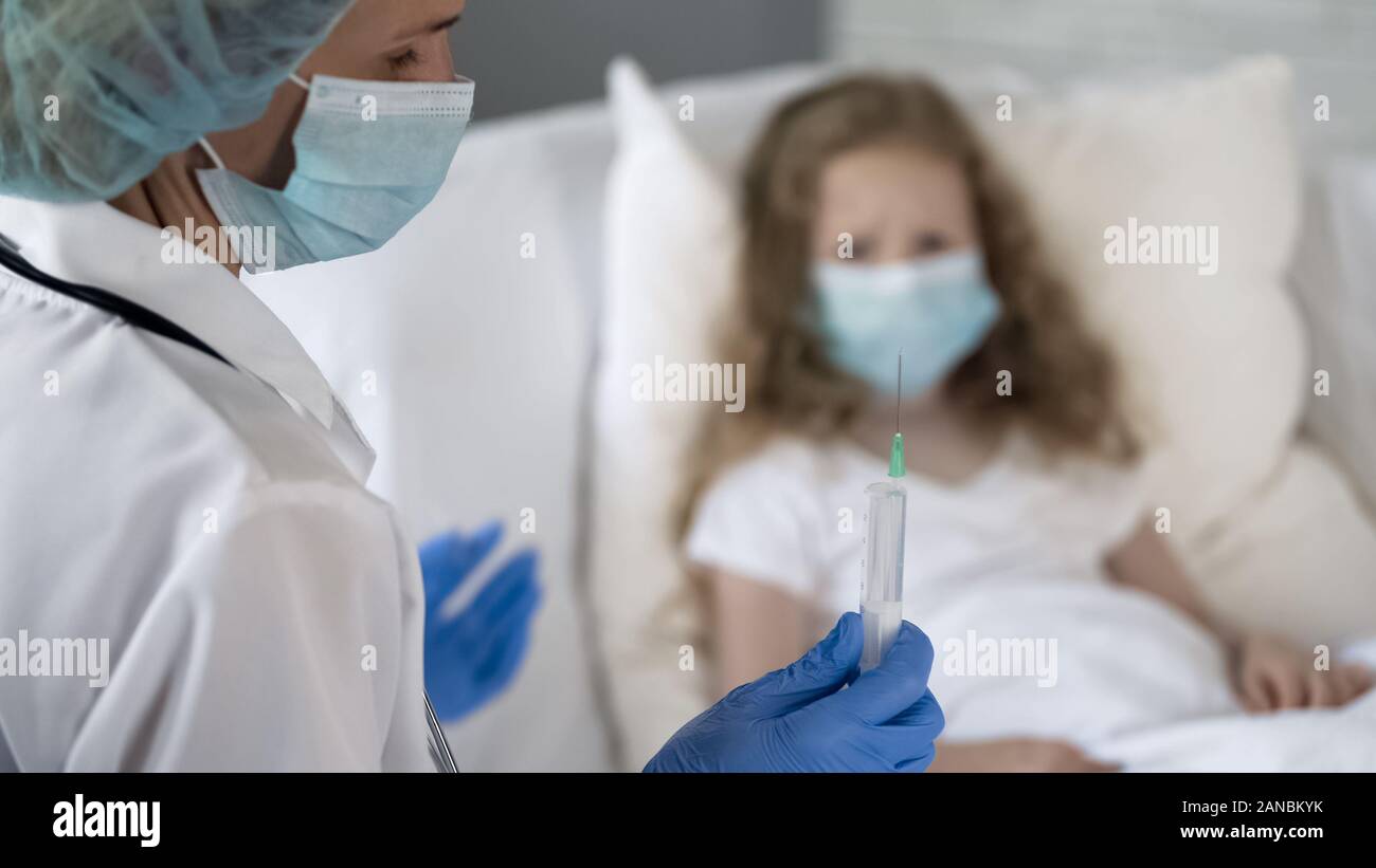 Unhappy scared child in medical face mask looking at nurse with syringe ...