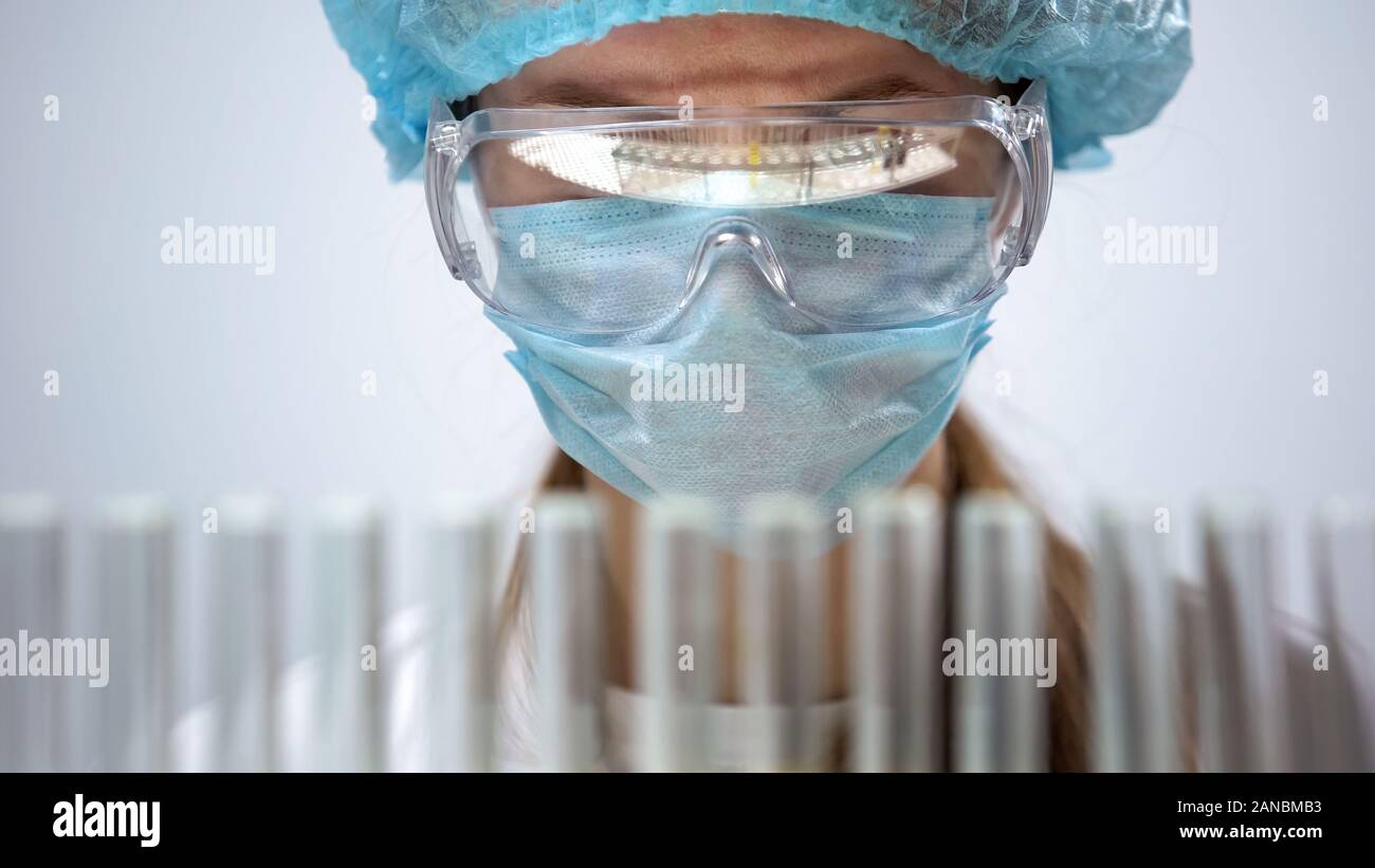 Female chemist in protective mask looking at test tubes, biological ...