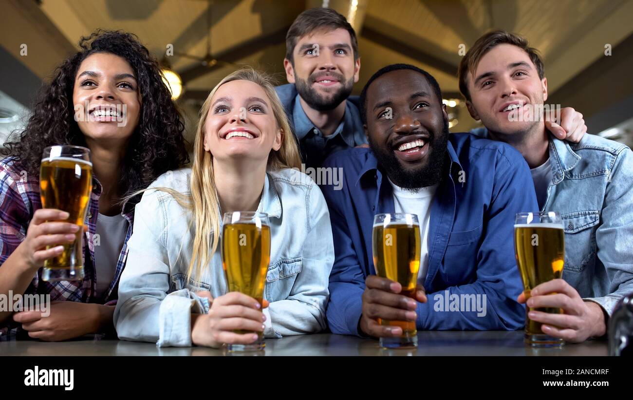 Multi-ethnic friends watching tv program on big screen in beer bar ...