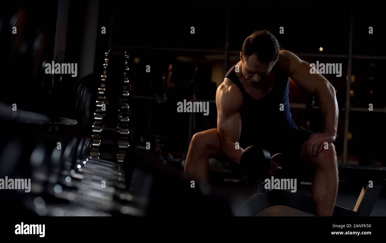 Strong-willed bodybuilder doing seated isolated dumbbell curl, evening ...