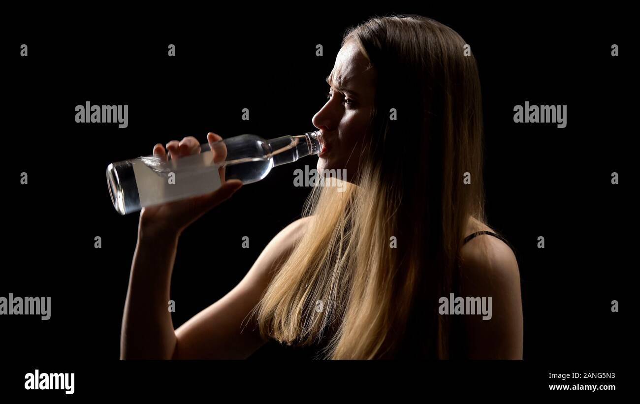 Young woman drinking alcohol from bottle against dark background ...