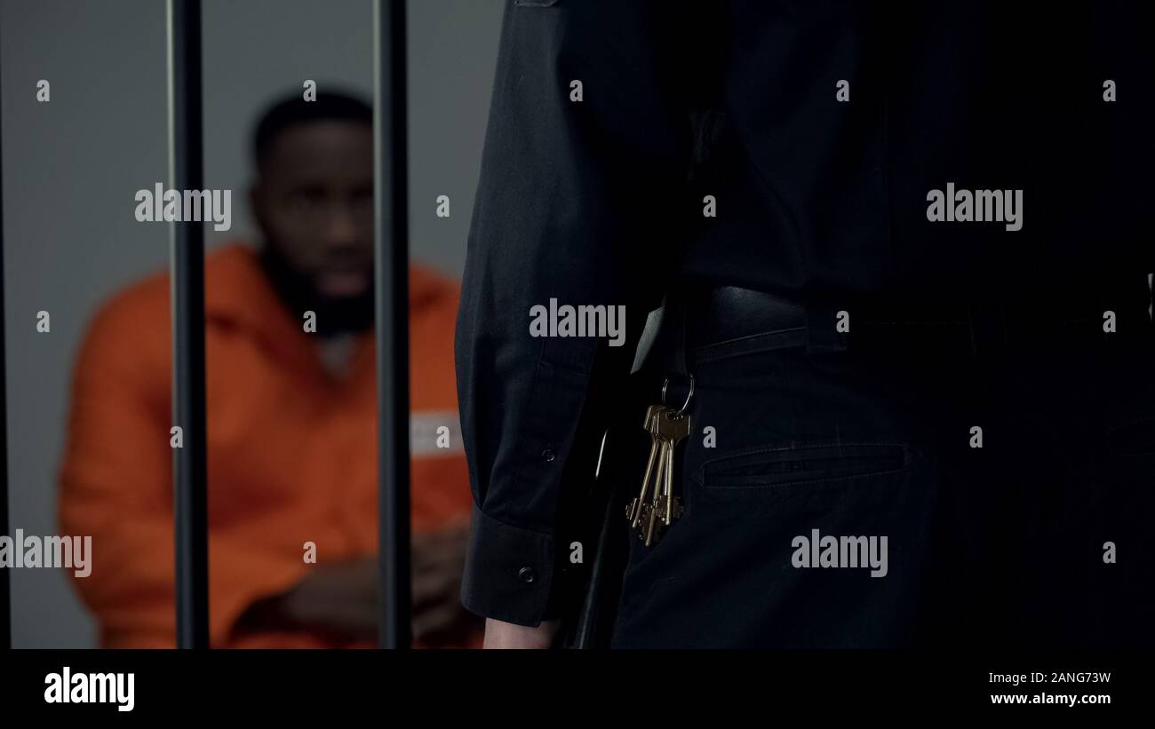 Security warden with keys looking at afro-american imprisoned male in ...