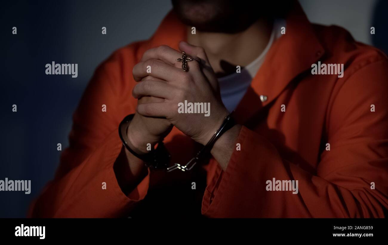 Imprisoned male in handcuffs holding cross and praying, feeling guilty ...