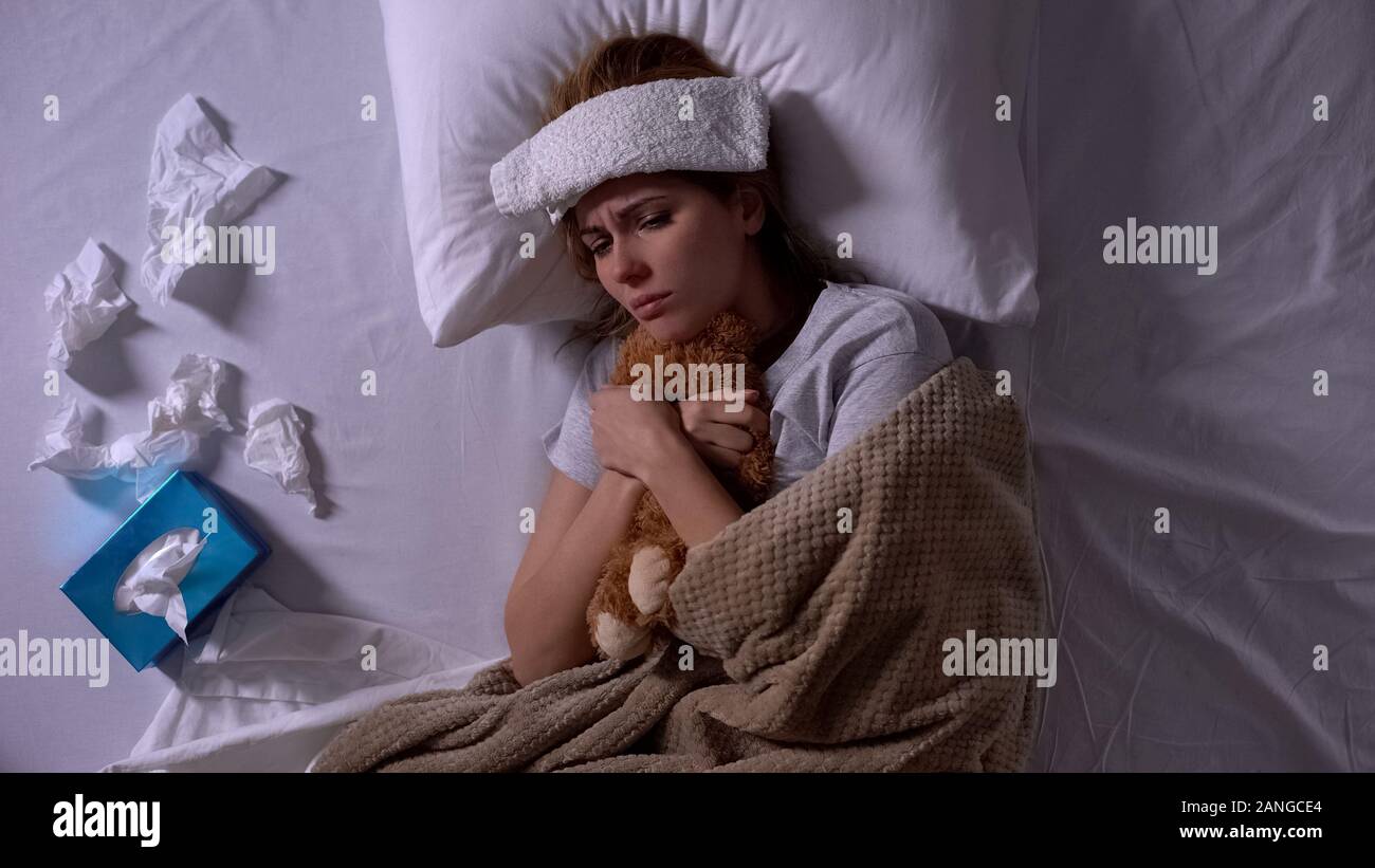 Lonely sick woman hugging teddy-bear lying in bed with compress and ...