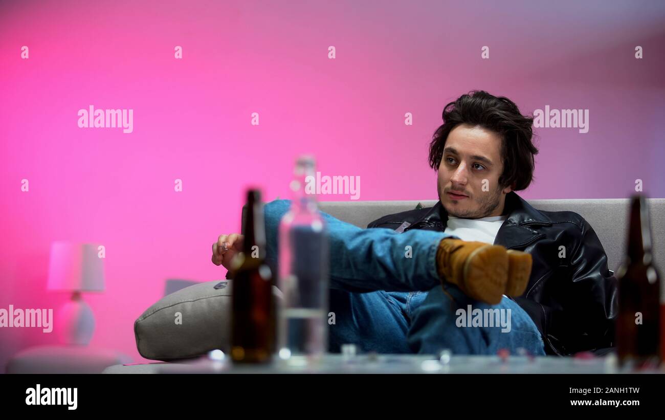 Intoxicated young man sitting on sofa, alcohol bottles on table, rave ...