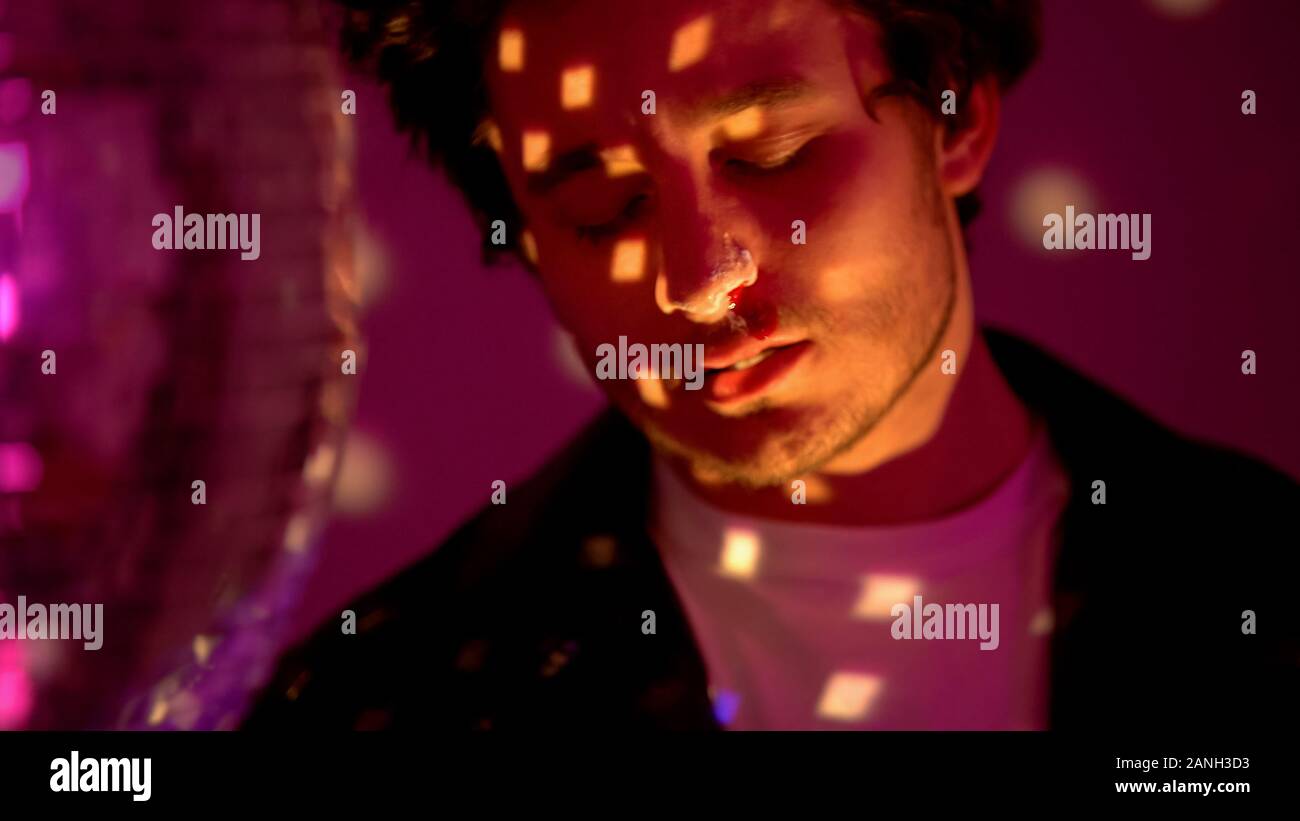 Addict man with bloody nose standing by disco ball, pill in mouth ...