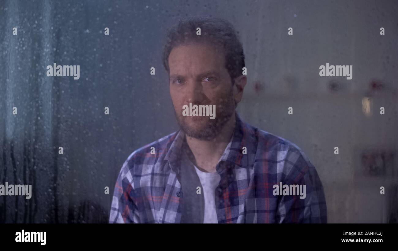 Lonely man sitting behind rainy window and looking at camera ...