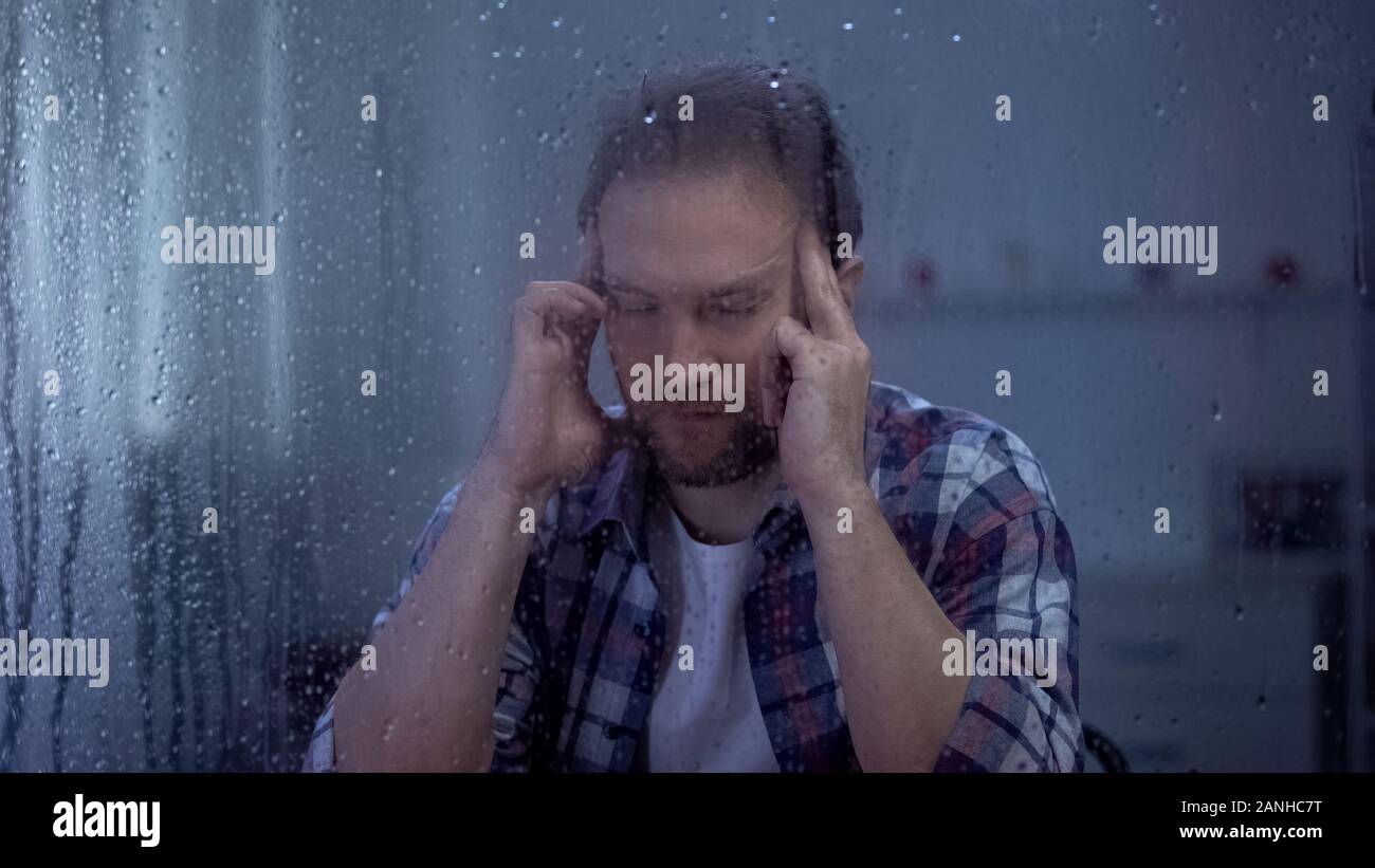Depressed man massaging temples behind rainy window, suffering from ...