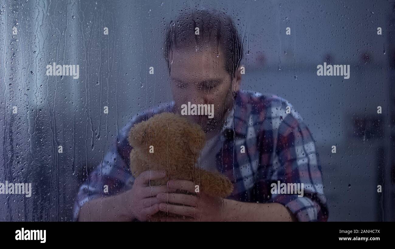 Lonely male holding teddy bear behind rainy window, missing child after ...