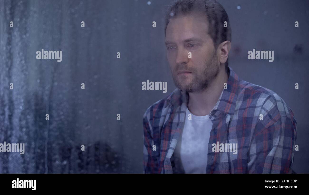 Sad man looking through window during rainy weather, thinking about ...