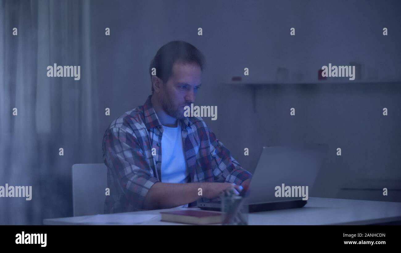 Focused middle-aged man working on laptop behind rainy window ...