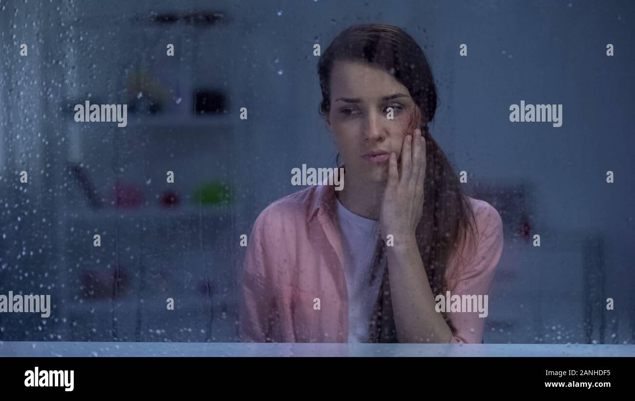 Depressed woman with wounded face sitting behind rainy window, assault ...