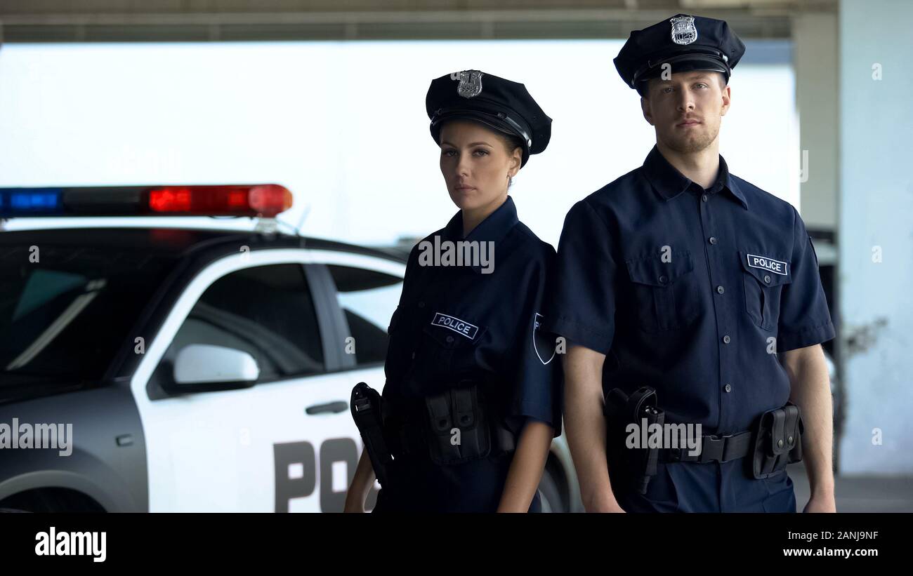Two serious policemen in uniform and service cap posing near patrol car ...