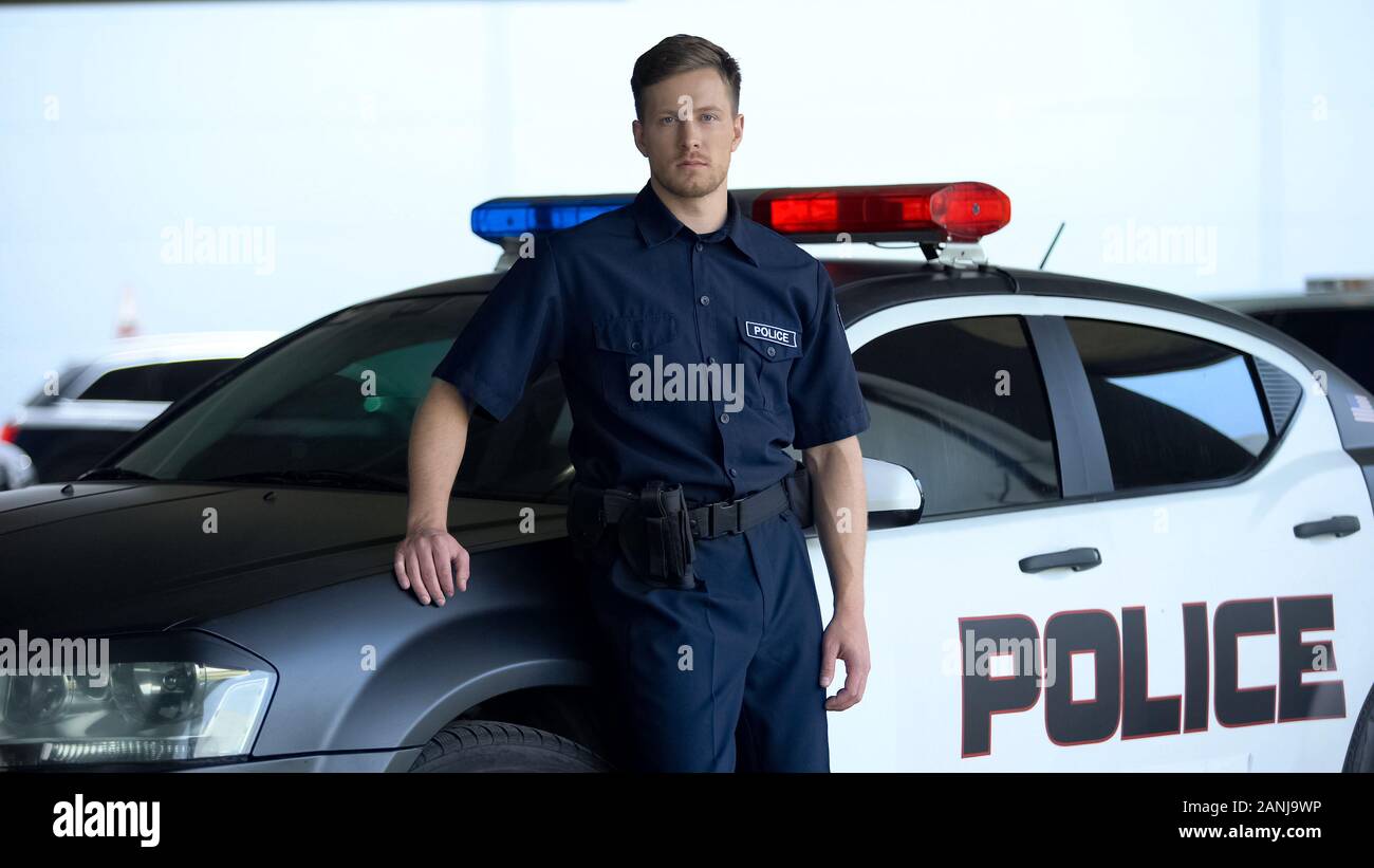 Serious policeman in uniform leaning on patrol car, maintenance of ...