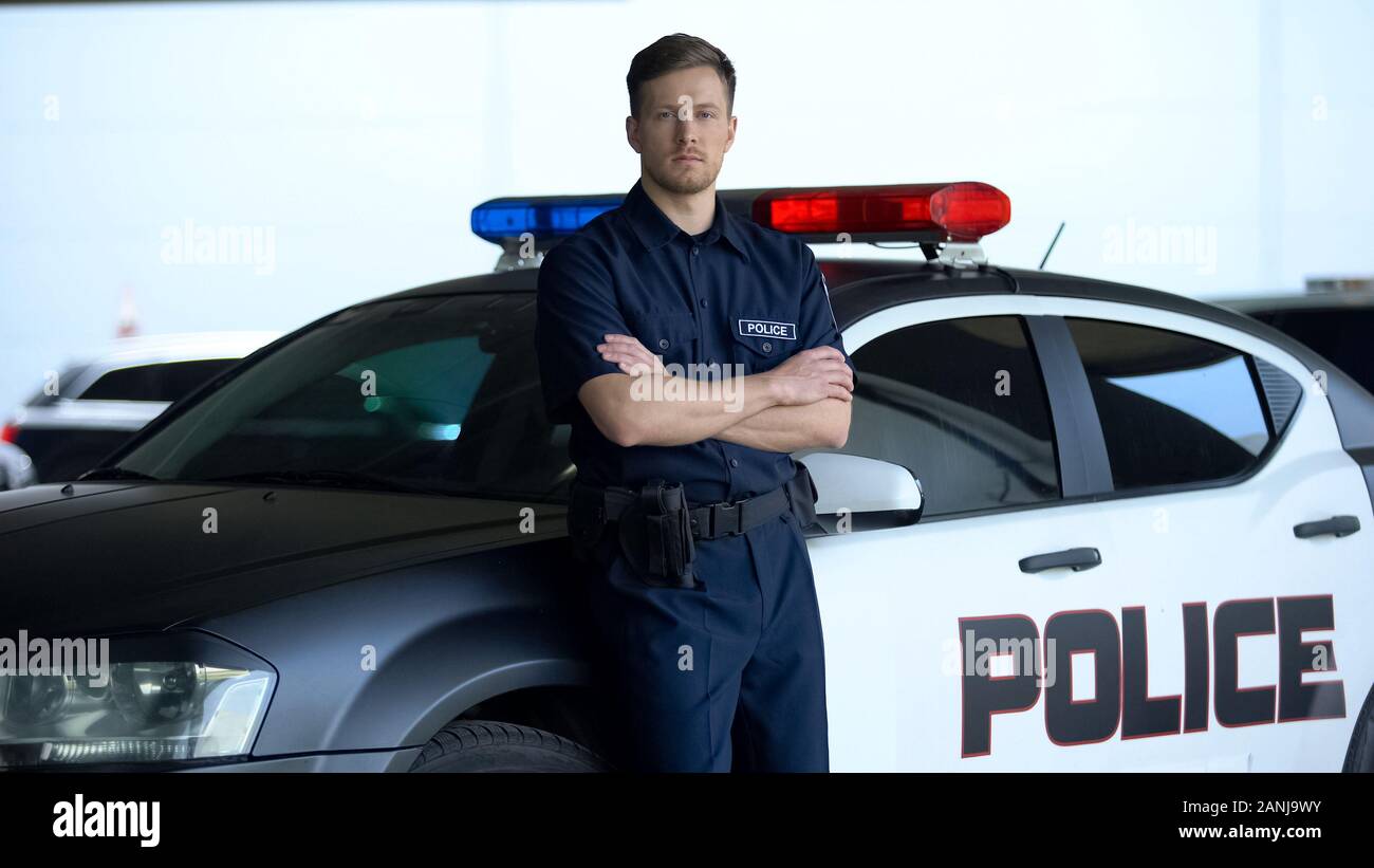 Professional policeman in uniform with crossed hands leaning on patrol ...