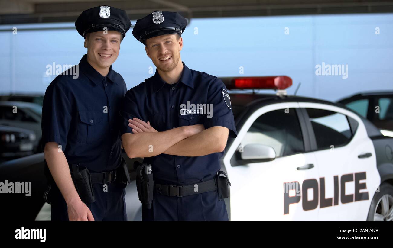 Positive police mates in hats smiling into camera against squad car ...