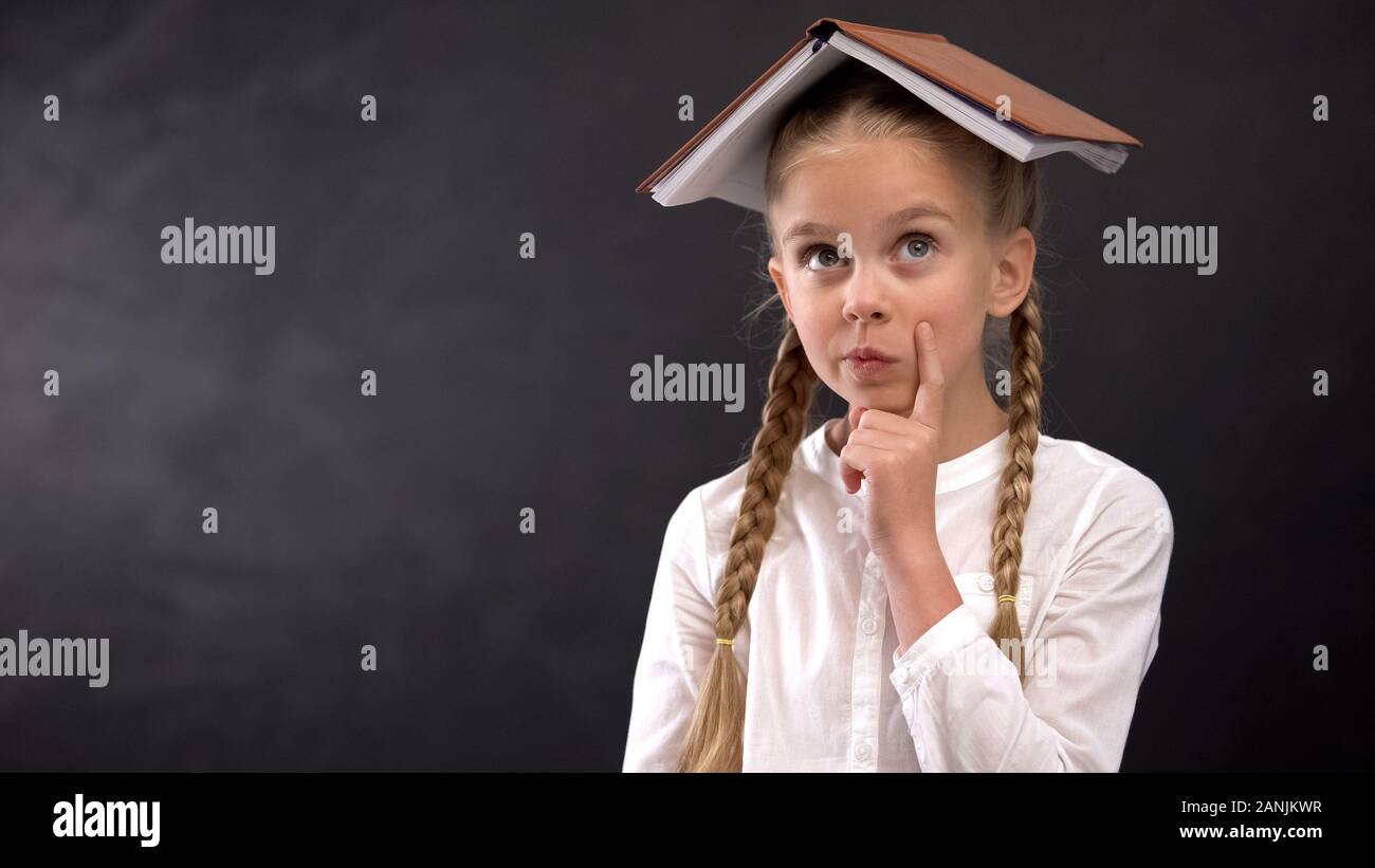Thoughtful pupil with book on head standing against blackboard ...