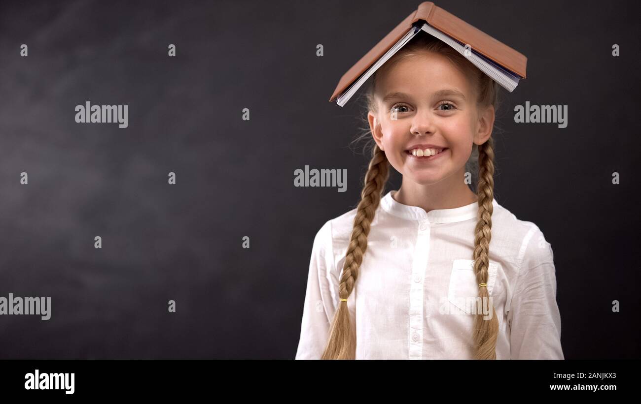 Smiling girl with book on head looking at camera, having fun on break ...