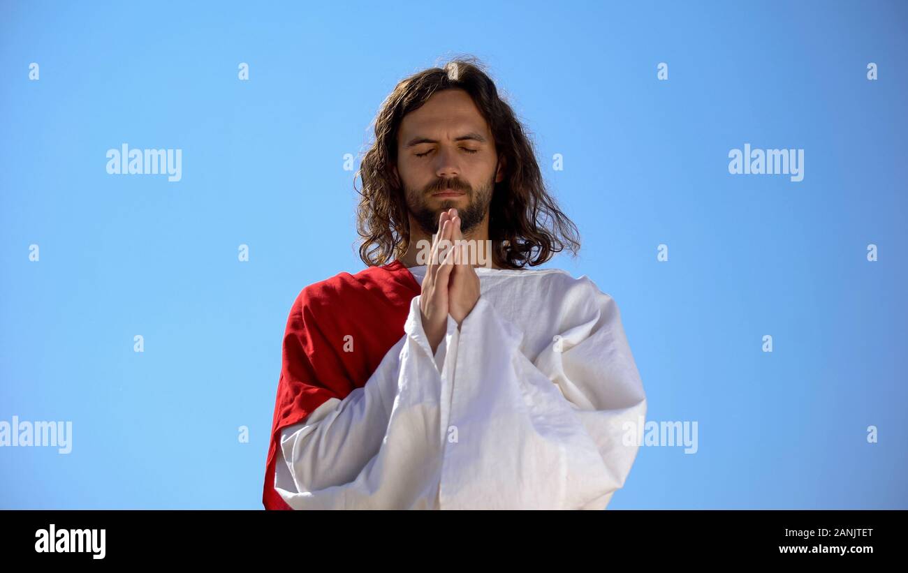 Jesus in robe praying against blue sky background, asking God to ...