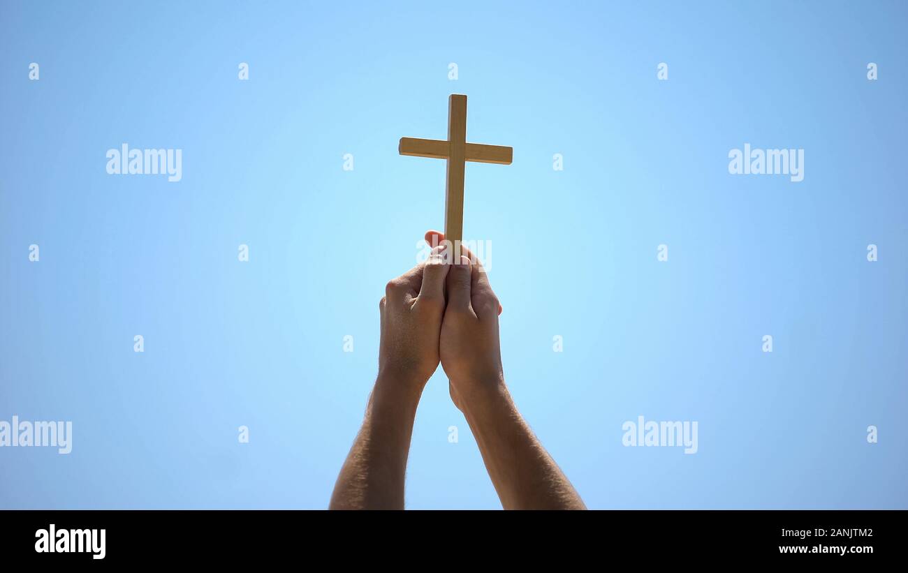 Male hands raising wooden cross to blue sky, religious conversion ...