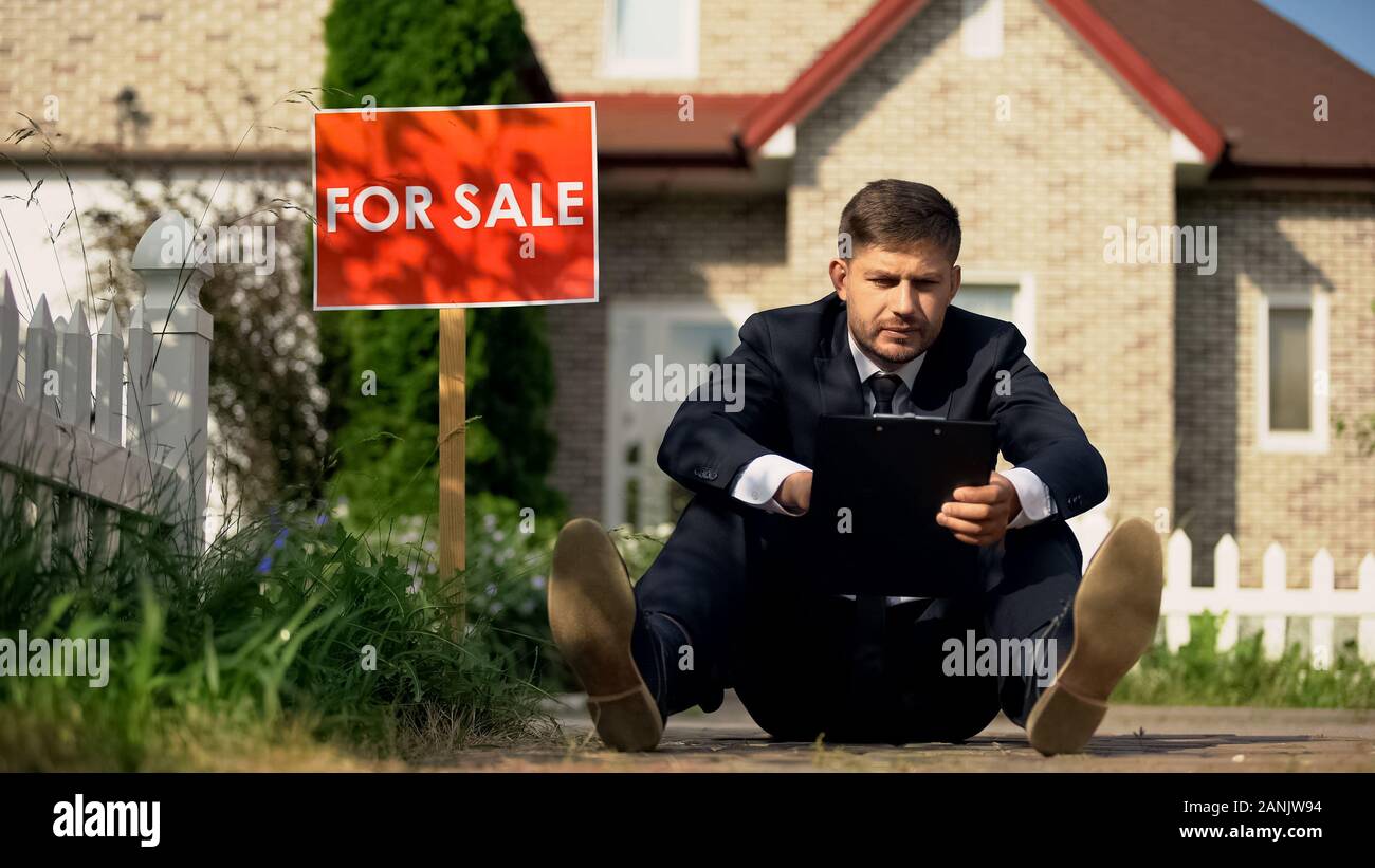 Tired estate broker sitting on ground near for sale signboard, failed ...