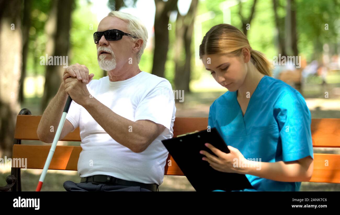Nurse taking notes sitting by blind old patient in park, walking with ...