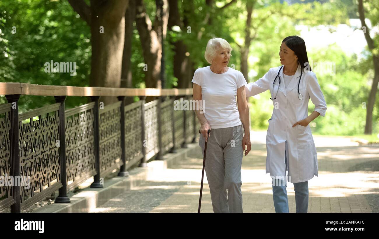 Doctor walking in park with elderly female patient with cane ...