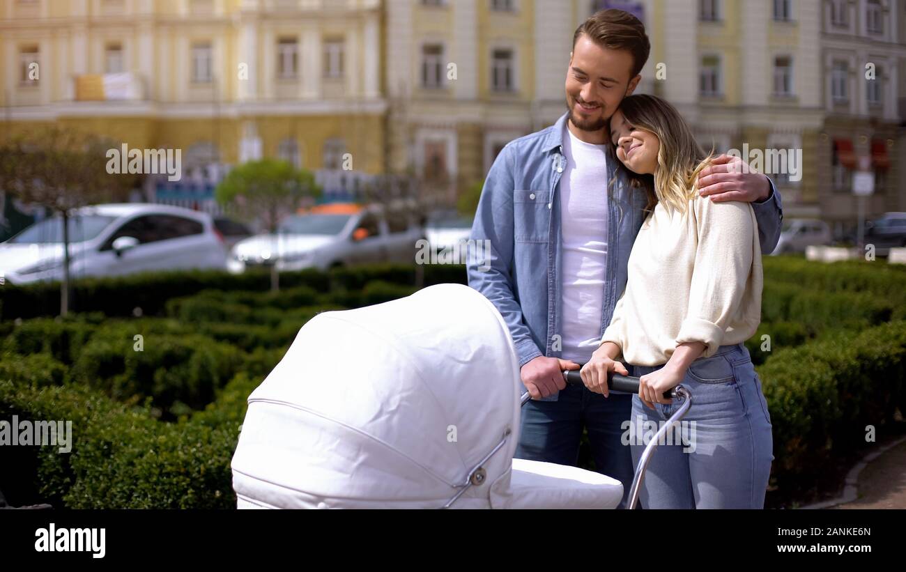 Smiling young parents walking in city garden pushing newborn pram ...