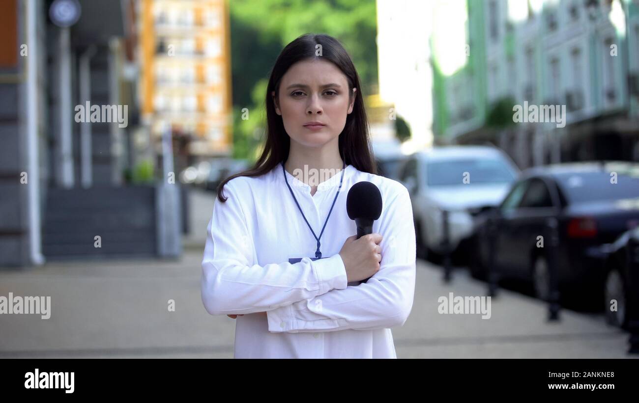 Serious female journalist with microphone and press badge looking at ...