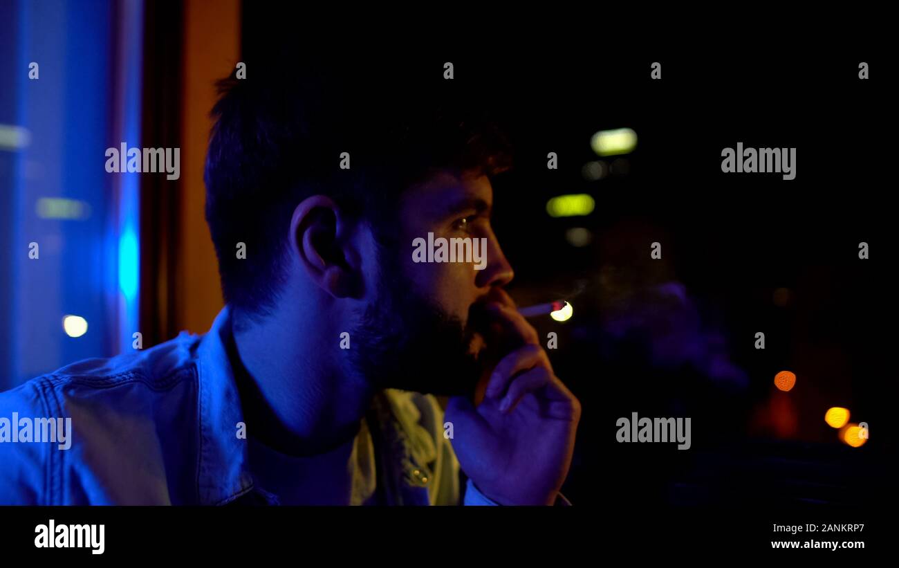 Young man smoking alone near window at night, worrying about life ...