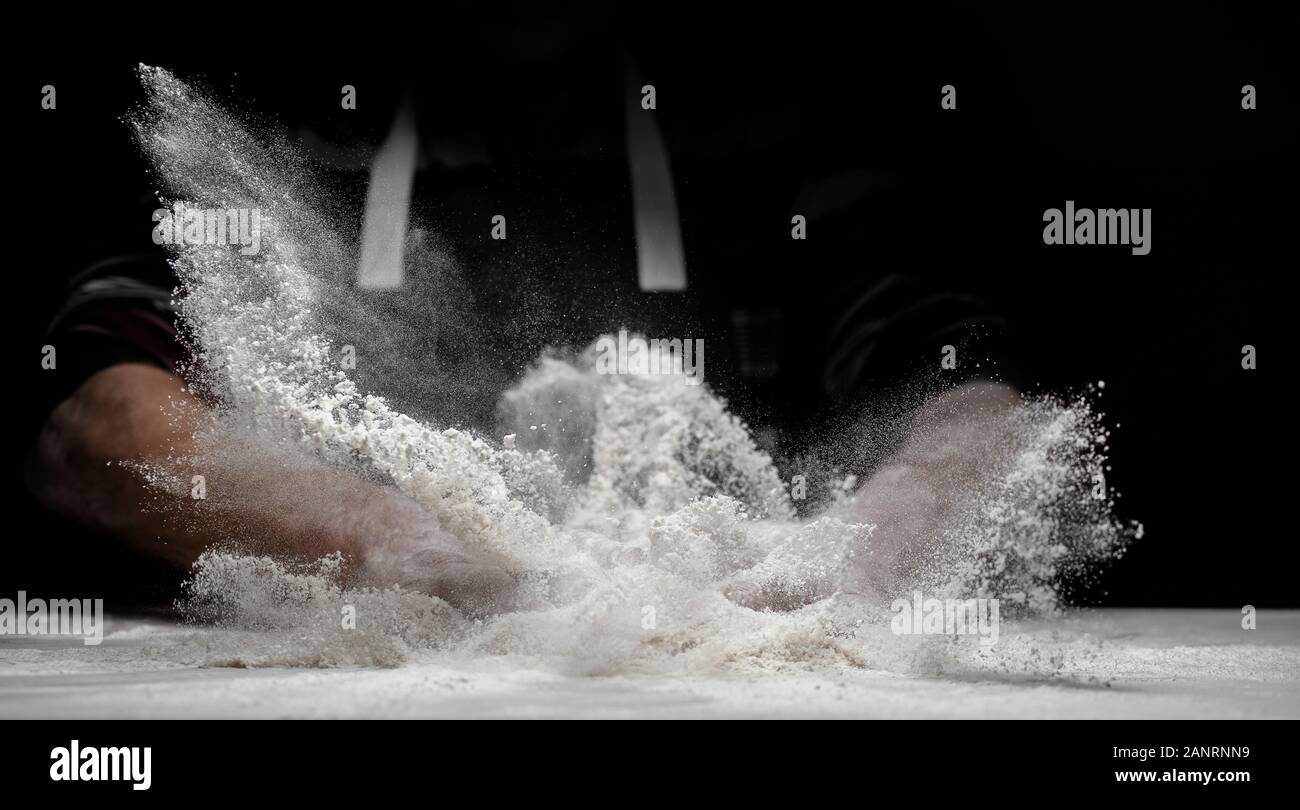 White flour flies in air on black background, pastry chef claps hands ...