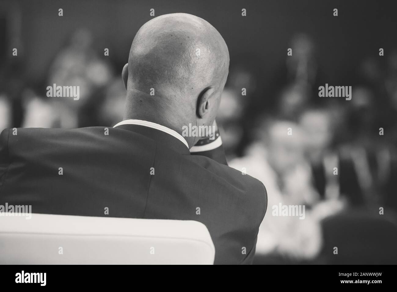 A back view of a male Speaker in a blue suit giving a talk on a Stage ...