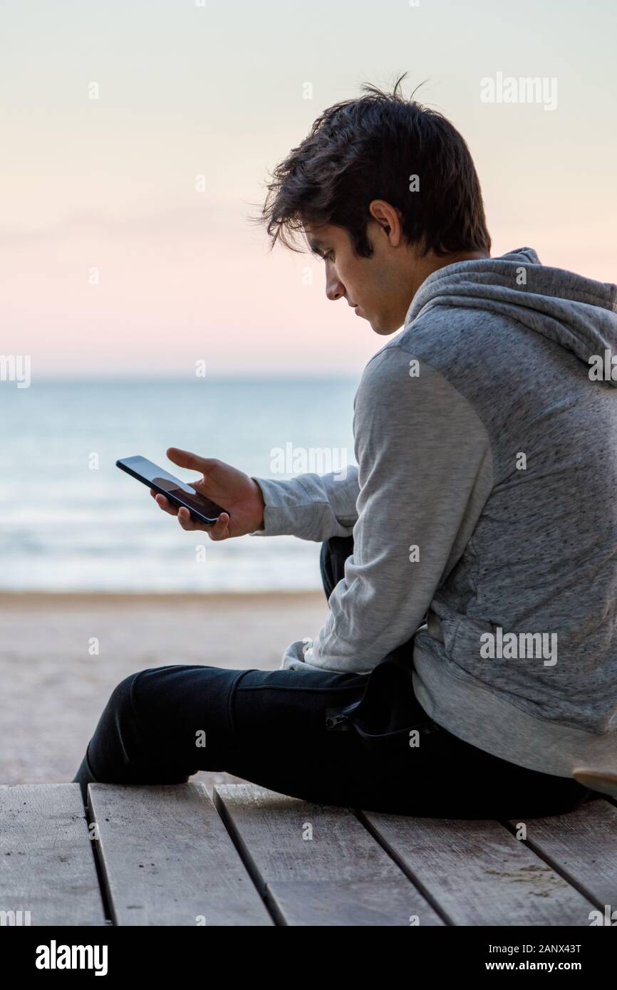 Young boy looking at his mobile phone on the beach next to his ...