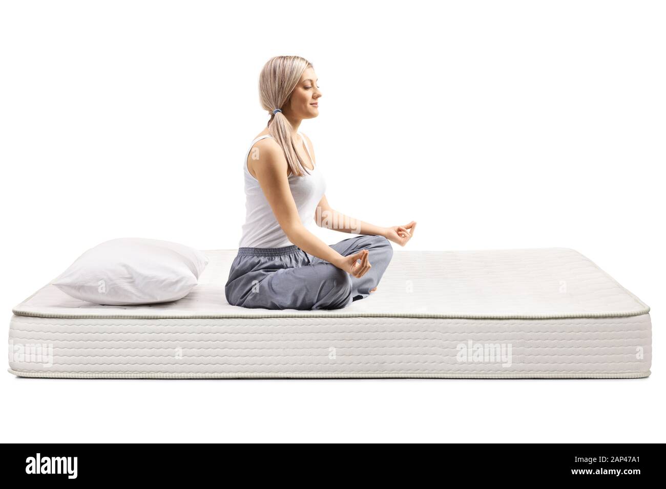 Young woman in pajamas sitting on a bed mattress in a meditation pose ...