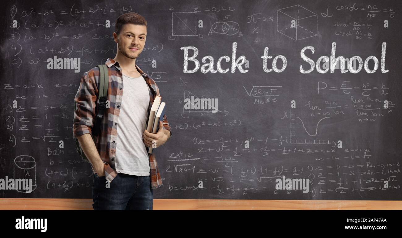 Male student with books standing in front of a school blackboard with ...