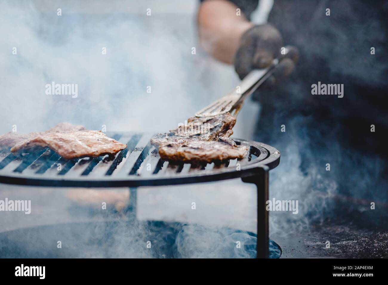 Cook grills steaks and pork ribs open fire. Turns meat over with tongs ...