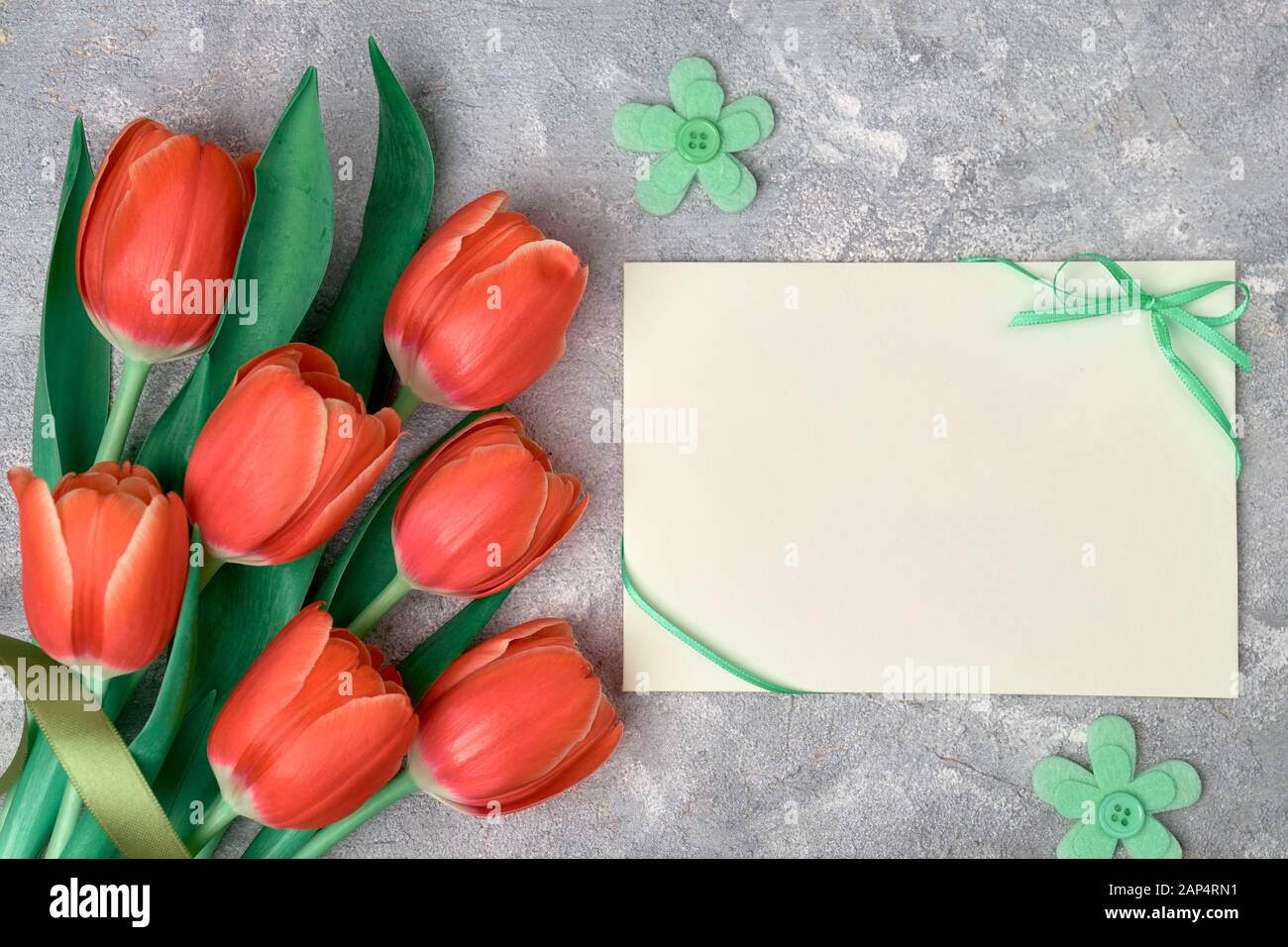 Spring greetings, flat lay with red tulips, green textile flowers and blank paper card on grey ...
