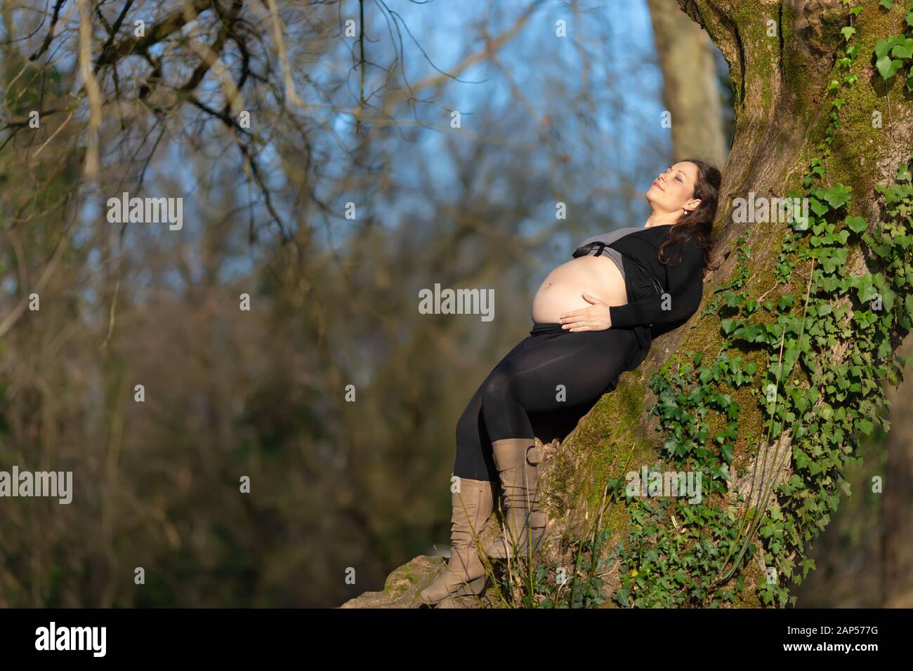 Pregnant woman leaning back on the tree in park, holding her bare belly ...