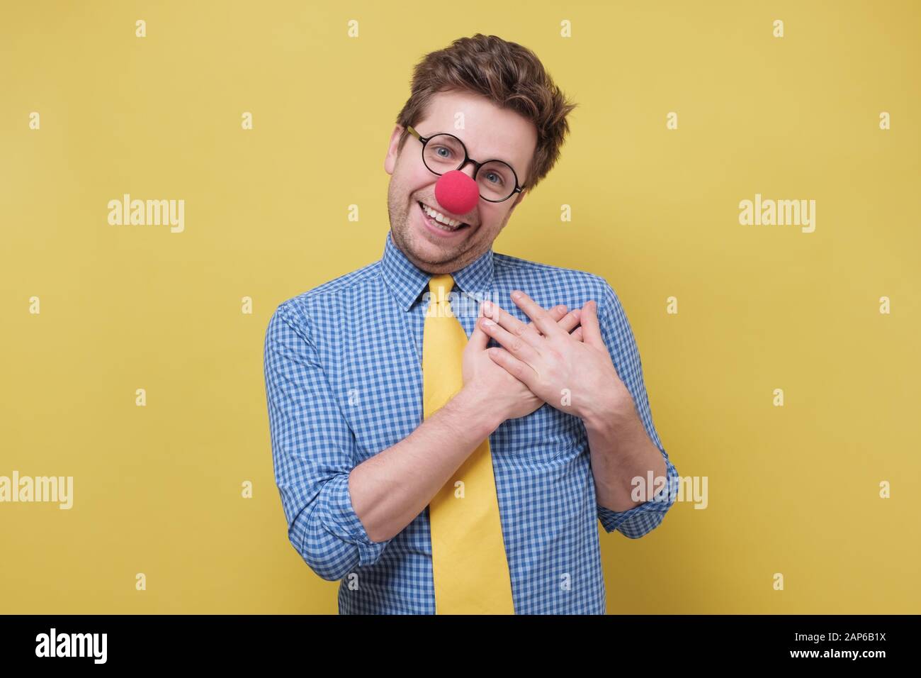 Cheerful clown man with red nose smiles happily, keeps hands on chest ...