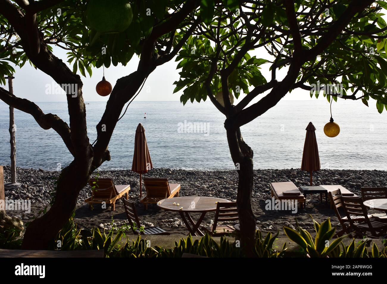 Volcanic pebble beach with wooden outdoor furniture under trees at calm ...