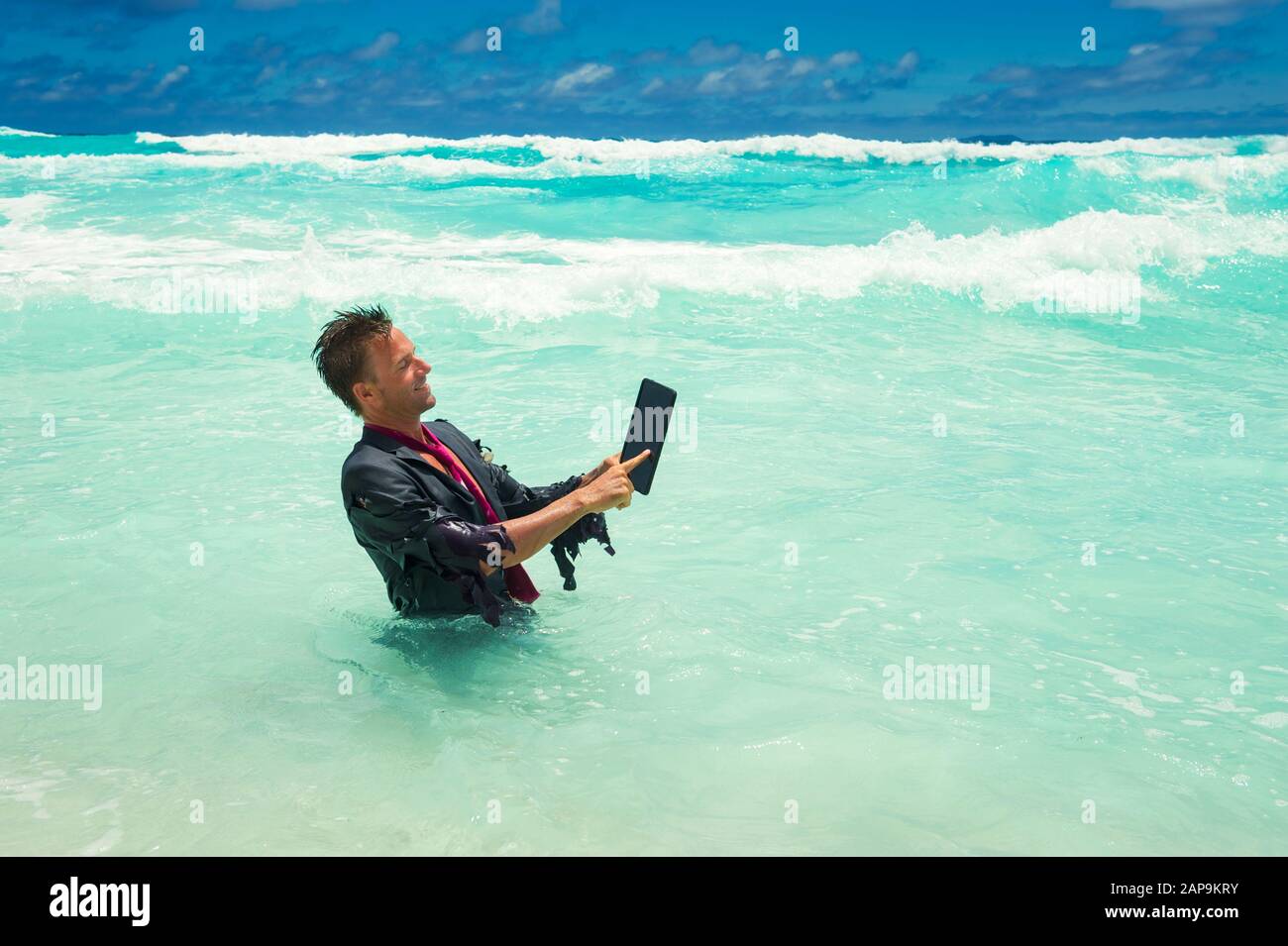 Cheerful castaway businessman using a tablet computer in tropical waves ...