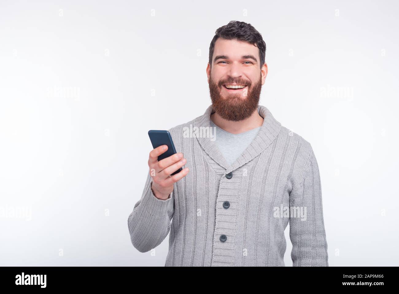 Happy man is holding his phone while smiling at the camera on white ...