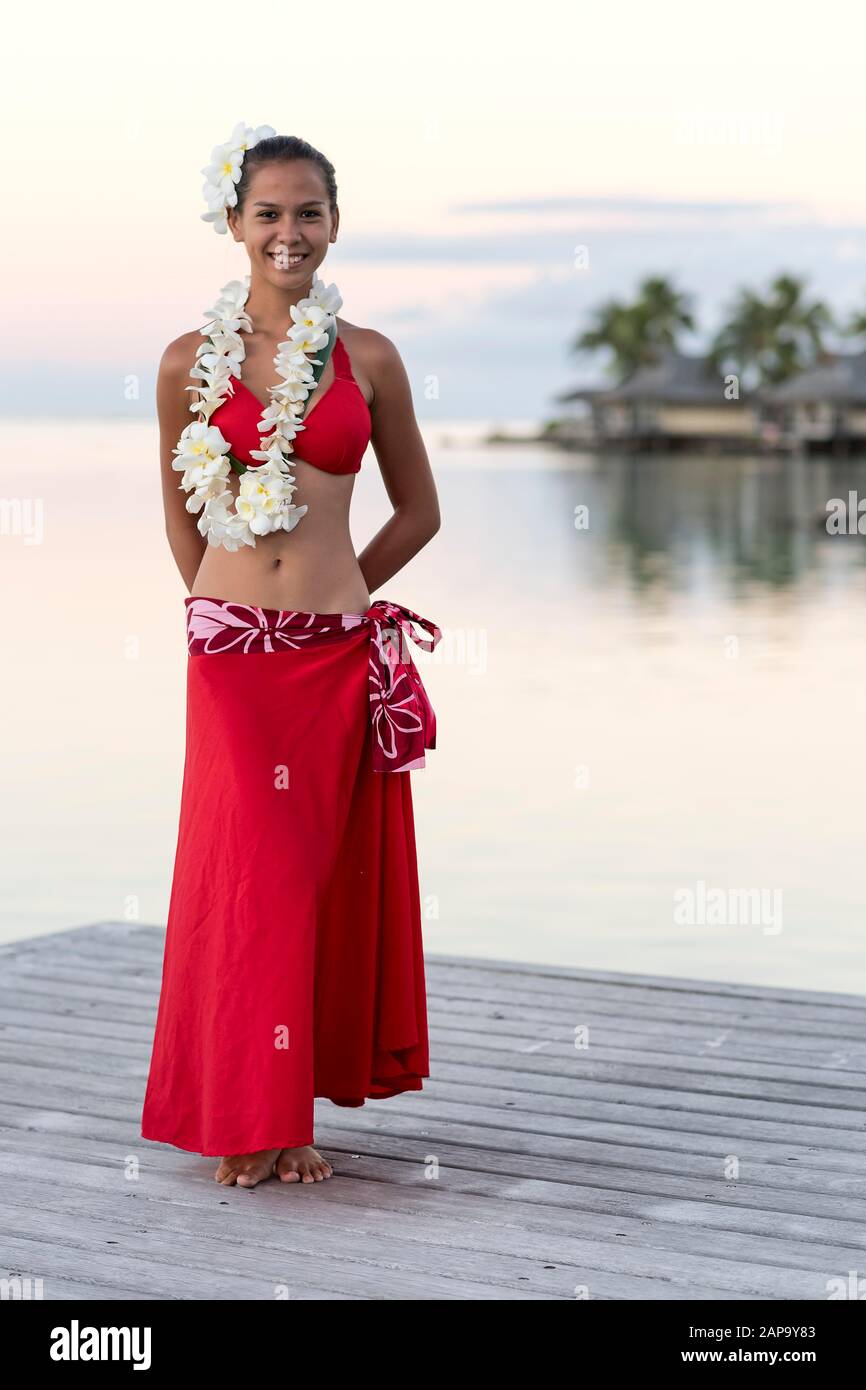 Native young woman with flower decoration stands on jetty by the water ...