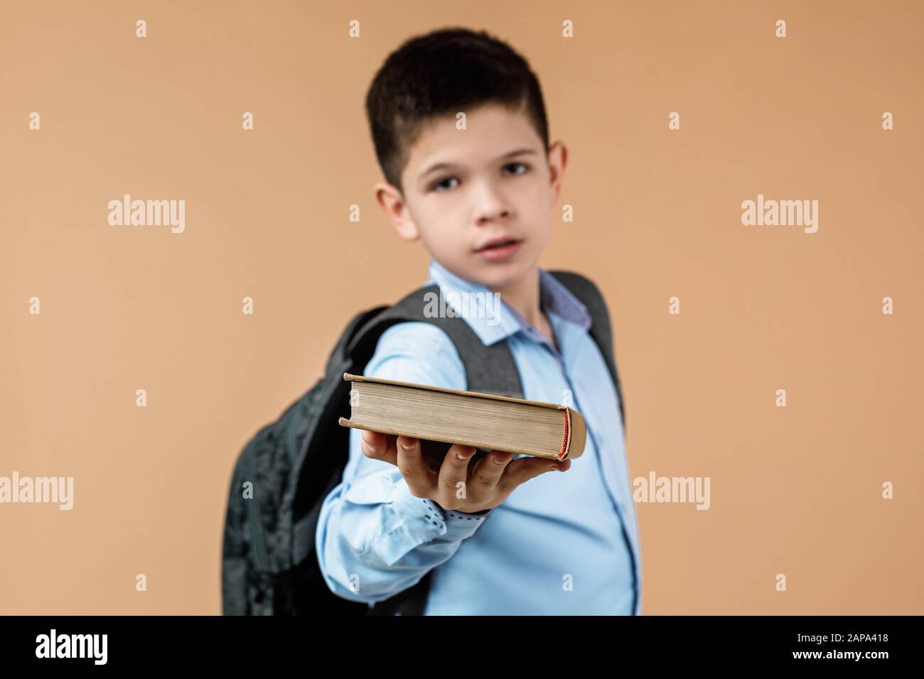 little cheerful school boy with backpack giving book over beige ...