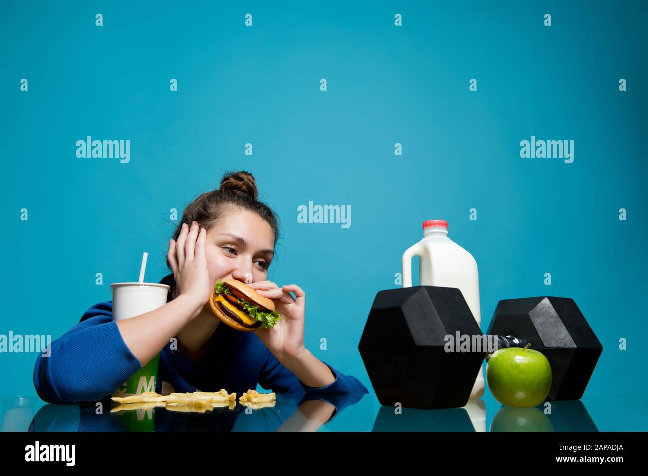 the girl lazily chews a burger and looks at the dumbbell, a bottle of ...